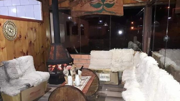 Uma área de estar interna aconchegante, com lareira a lenha, sofás brancos macios e uma parede de madeira rústica.