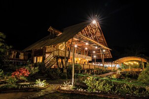 Uma casa de madeira grande com vários níveis e uma área externa coberta, cercada por jardins bem cuidados.