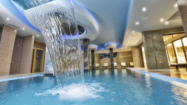 Piscina interior moderna com uma cascata central, rodeada por áreas de descanso e um teto alto com iluminação embutida.
