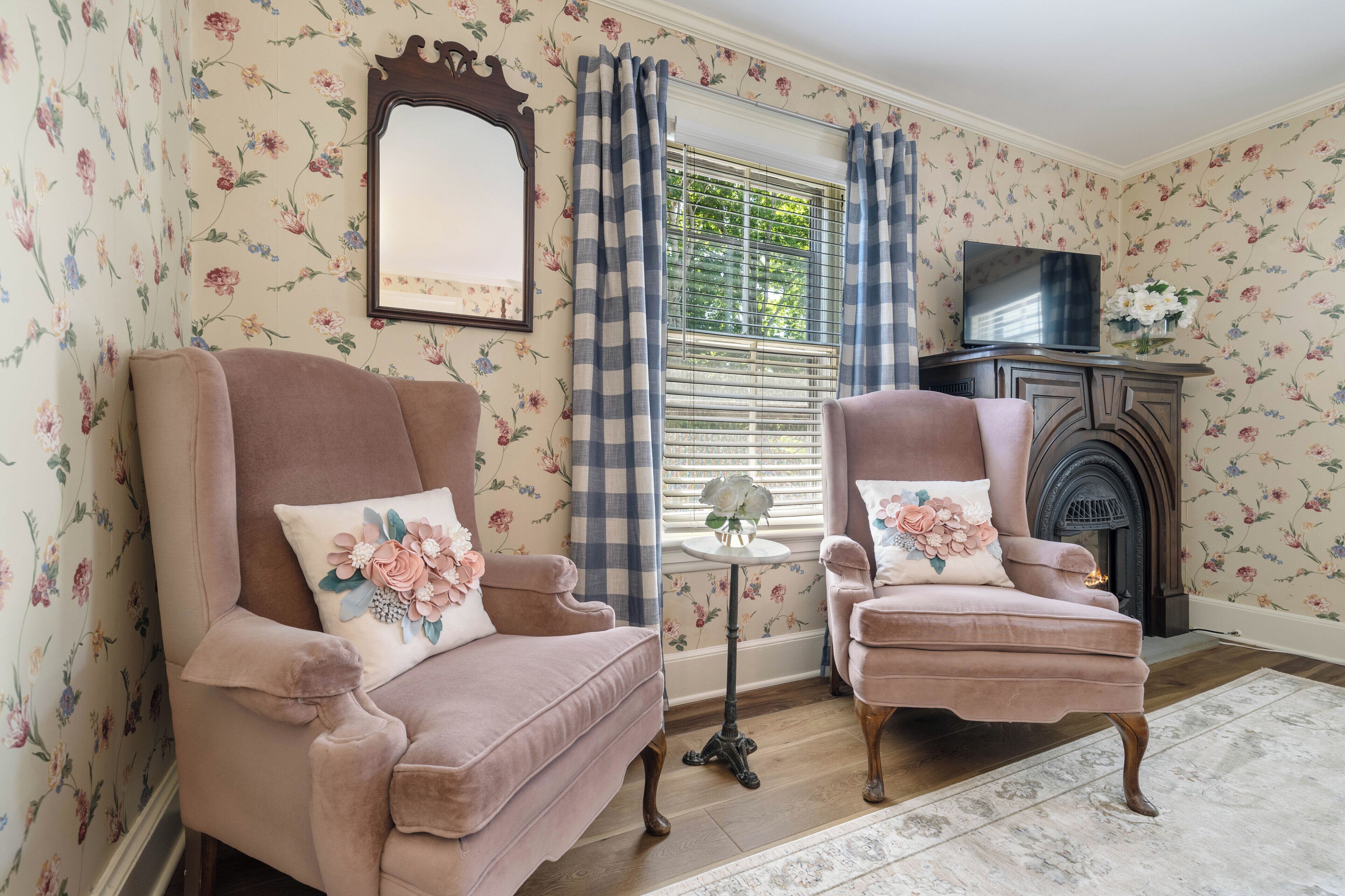 Quarto com papel de parede floral, dois poltronas com almofadas estampadas, uma mesa pequena com um vaso, uma televisão e uma lareira.