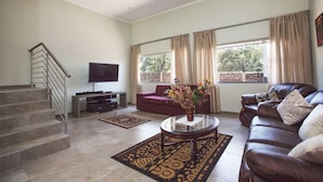 Uma sala de estar com escada, televisão de tela plana, sofá, poltrona de couro e mesa de centro com um vaso de flores.