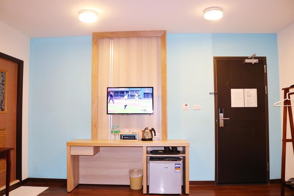 Quarto com escrivaninha de madeira, televisão na parede, mini-geladeira e porta com um aviso.