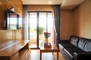 Uma sala de estar com um móvel de madeira para televisão, um sofá de couro preto, uma mesa de centro de madeira com um vaso de flores e uma porta de vidro deslizante para uma varanda externa.