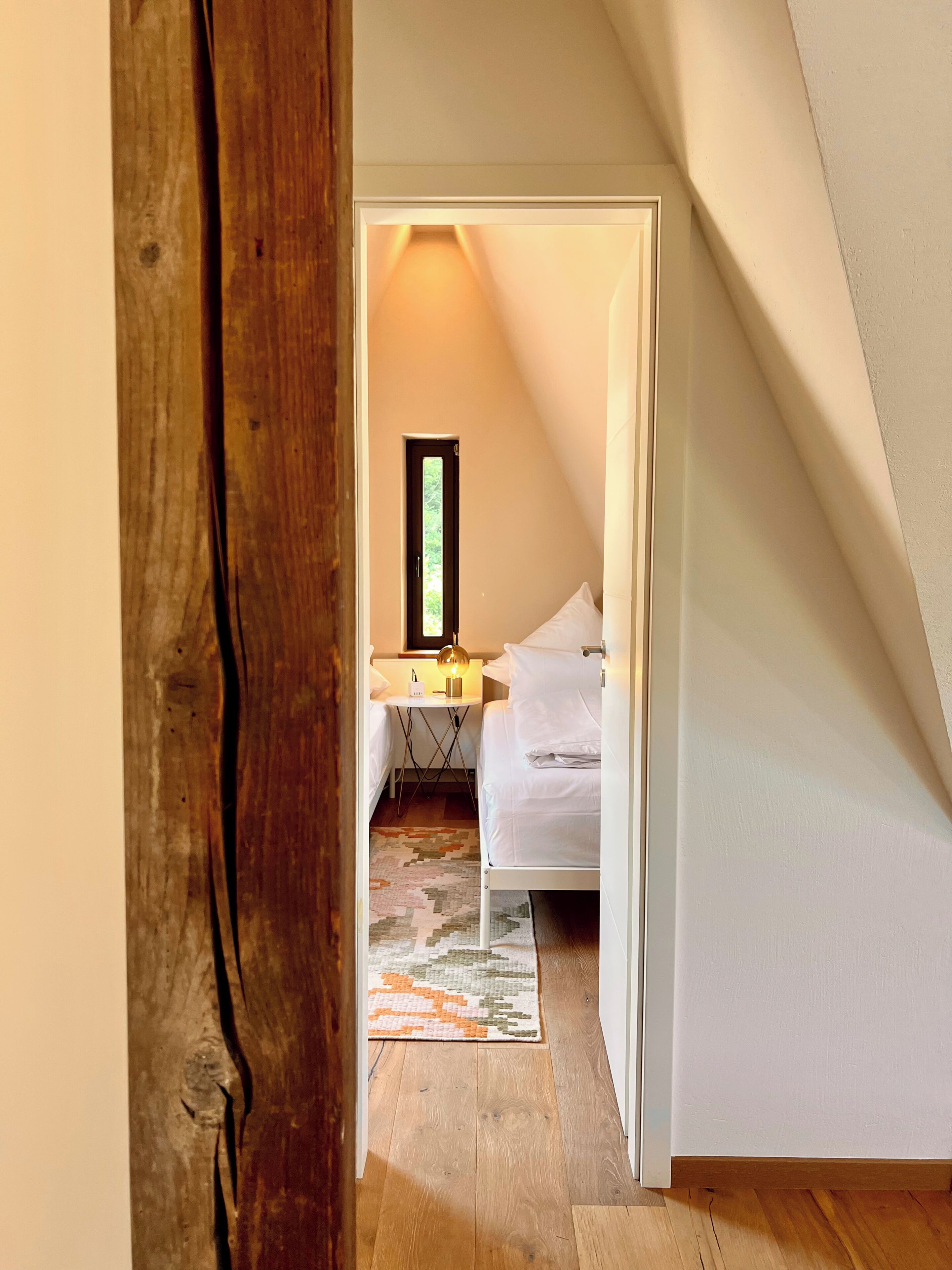 Um quarto com uma cama, uma mesa de cabeceira com abajur, uma janela e uma porta que dá para outro cômodo.
