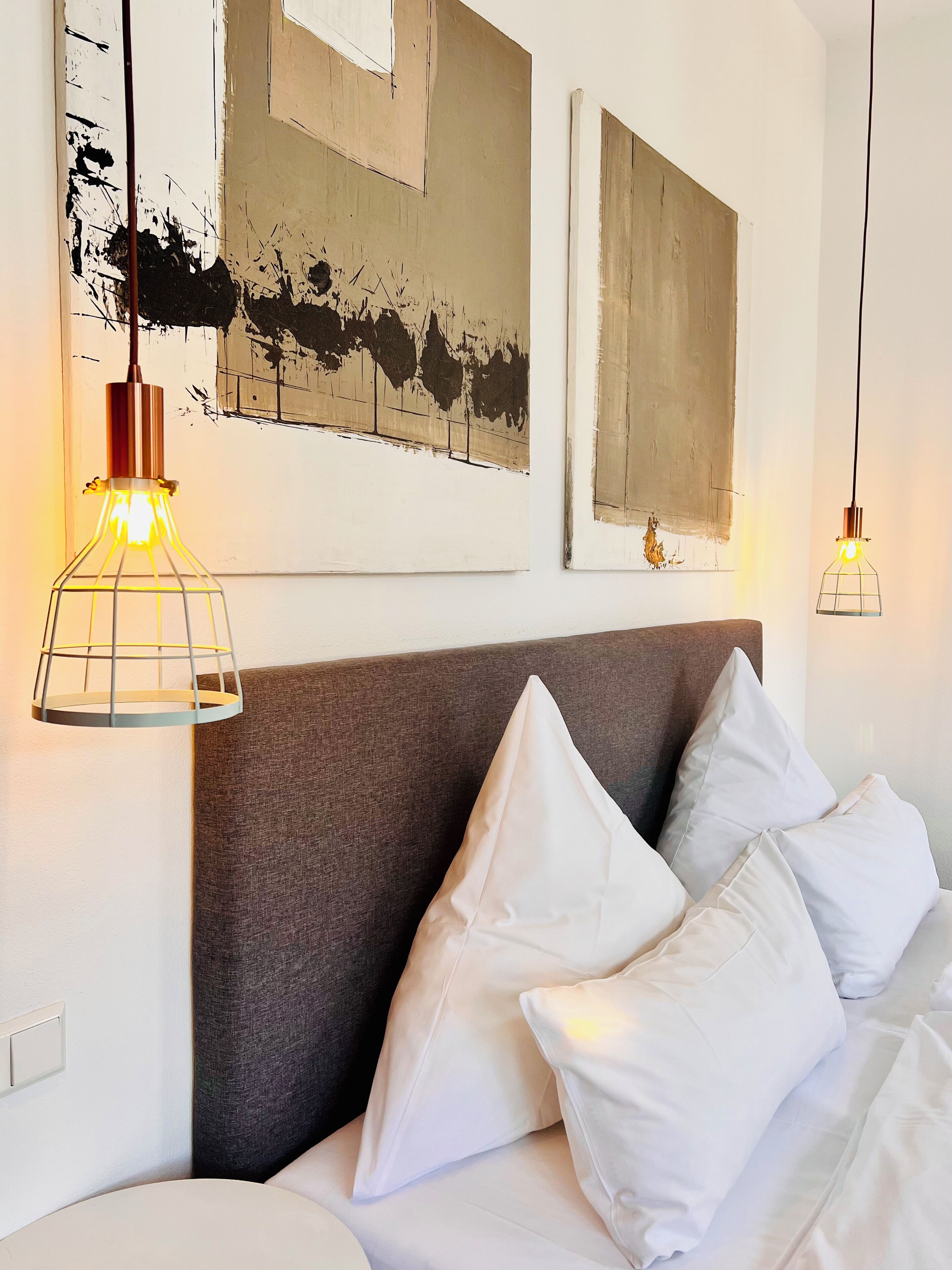 Uma cama bem arrumada com almofadas brancas e cabeceira cinza, duas luminárias pendentes com lâmpadas aparentes e duas pinturas abstratas na parede.