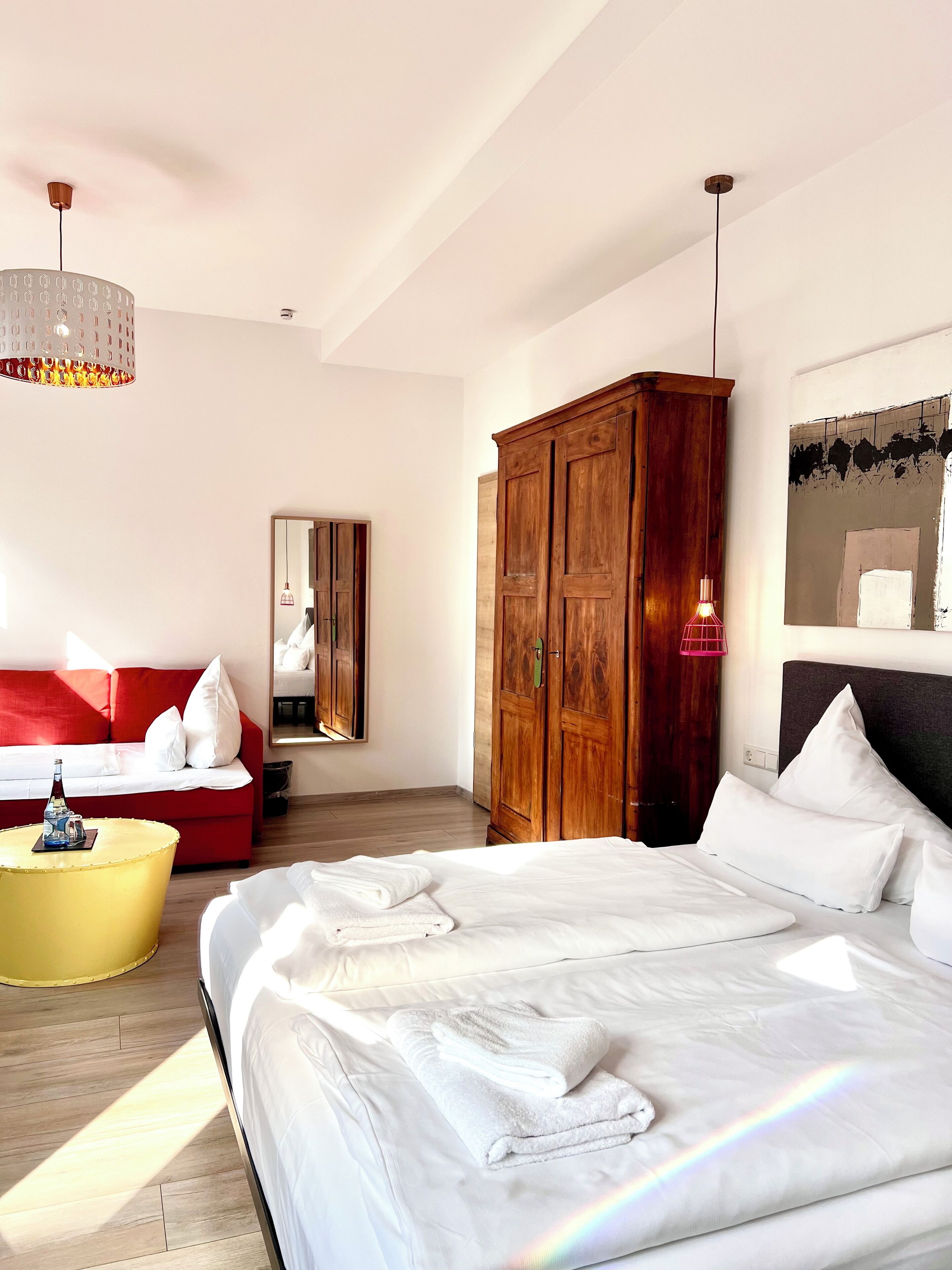 Quarto de hotel moderno com uma cama grande, um sofá vermelho, um guarda-roupa de madeira e um banquinho amarelo.