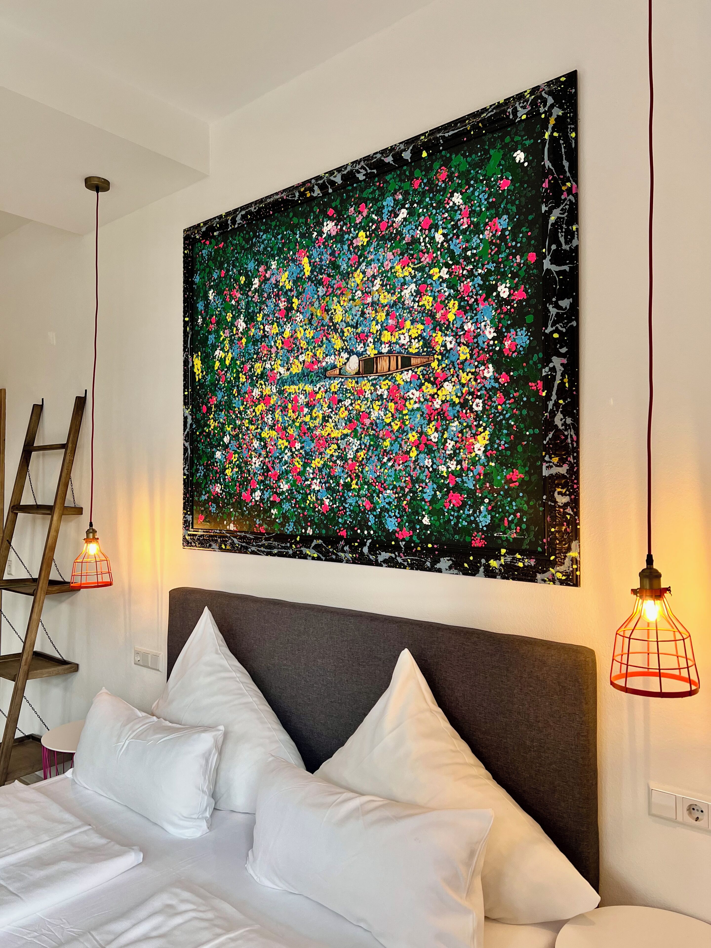 Quarto com cama, almofadas, uma escada e uma pintura abstrata e colorida na parede.