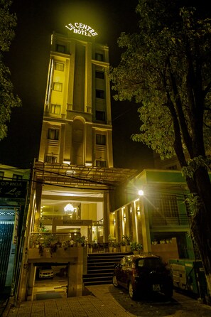 Um hotel com uma placa que diz "Le Centre Hotel" no topo. O edifício tem vários andares com janelas e varandas visíveis. Há plantas em vasos no andar térreo e um carro estacionado na frente.