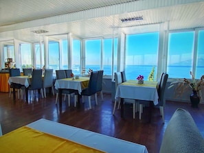 Restaurante com mesas e cadeiras dispostas para refeições, grandes janelas com vista para o mar e teto com ventilação.
