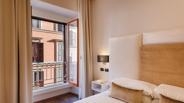 Quarto de hotel com uma cama grande, uma mesinha de cabeceira, uma luminária na parede e uma janela com vista para edifícios.