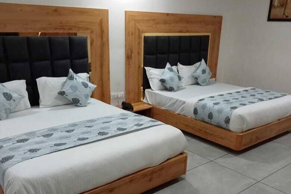 Quarto de hotel com duas camas, cada uma com estrutura de madeira e cabeceira escura, roupa de cama branca e manta estampada.