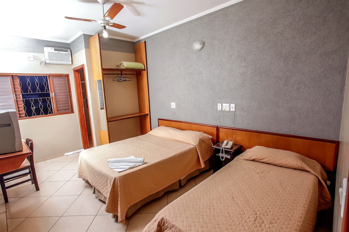 Quarto de hotel com duas camas, uma escrivaninha, uma cadeira, um ventilador de teto e um aparelho de ar condicionado.