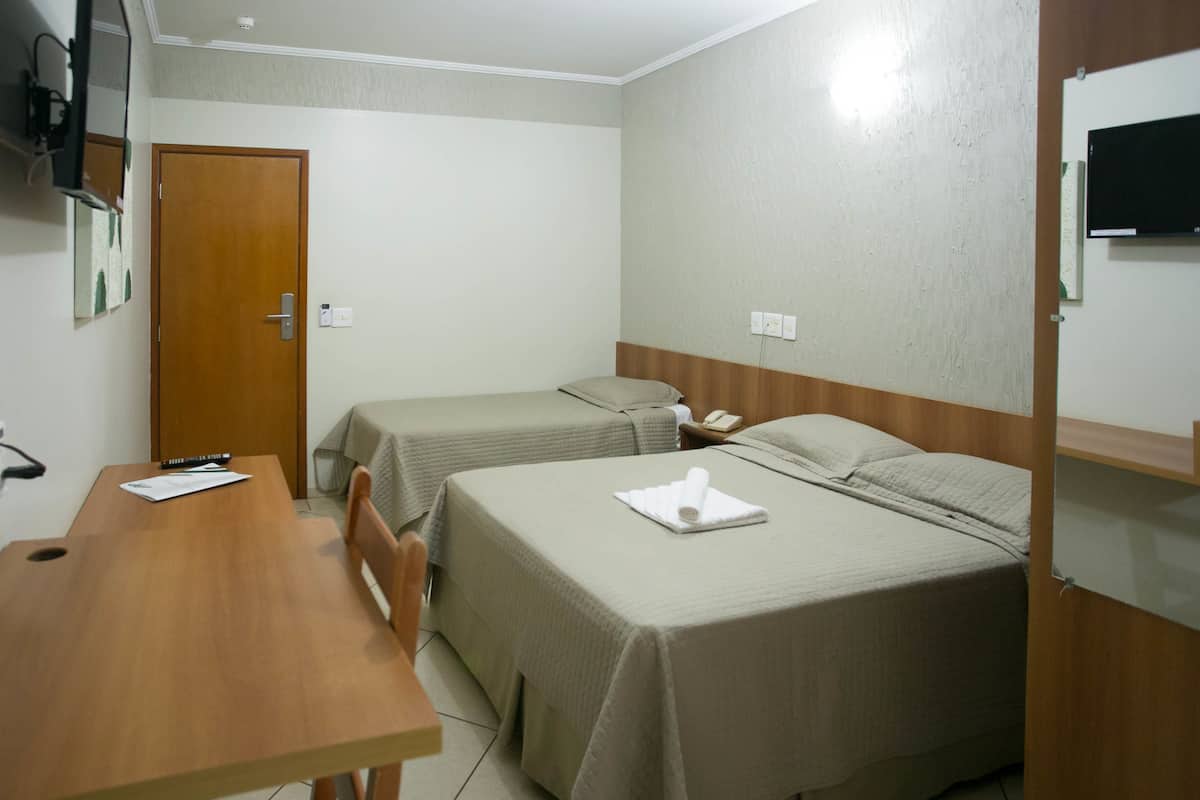 Quarto de hotel com duas camas de solteiro, uma escrivaninha de madeira, uma cadeira, um telefone e uma televisão na parede.