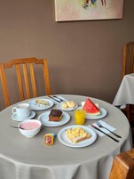 Uma mesa redonda com toalha branca, diversos itens de pequeno-almoço, incluindo uma fatia de melancia, uma tigela de cereais e uma torrada.
