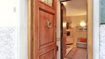 Uma porta de madeira com puxador de latão, dando entrada a um quarto com um tapete estampado, um sofá e uma prateleira fixada na parede com objetos decorativos.