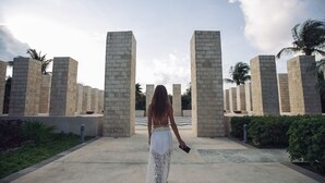 Uma mulher caminhando por um pátio com pilares de pedra e palmeiras.