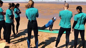 Um grupo de pessoas em fatos de mergulho numa praia, algumas em pé e outras num prancha de surf, com o oceano e um céu limpo ao fundo.
