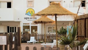 Um hostel com uma placa que diz "Sagres Sun Stay Hostel and Surf Camp". Há cercas de madeira, um guarda-sol de palha e uma espreguiçadeira à vista.