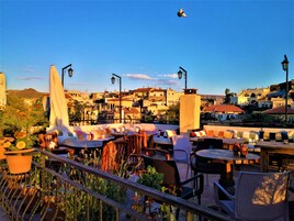 Uma área externa com mesas e cadeiras, plantas em vasos e vista para um horizonte urbano com edifícios e uma montanha ao fundo.