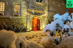 Um edifício histórico com entrada imponente, arco ricamente decorado e um tapete vermelho que convida a entrar. Neve cobre o chão e a vegetação ao redor da entrada.