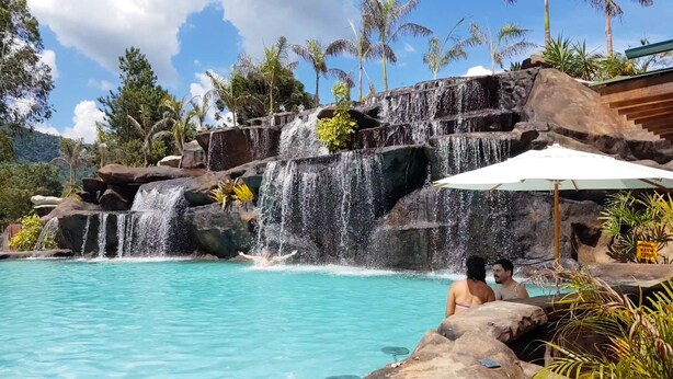 Piscina com cachoeira, rodeada por vegetação tropical e céu claro.