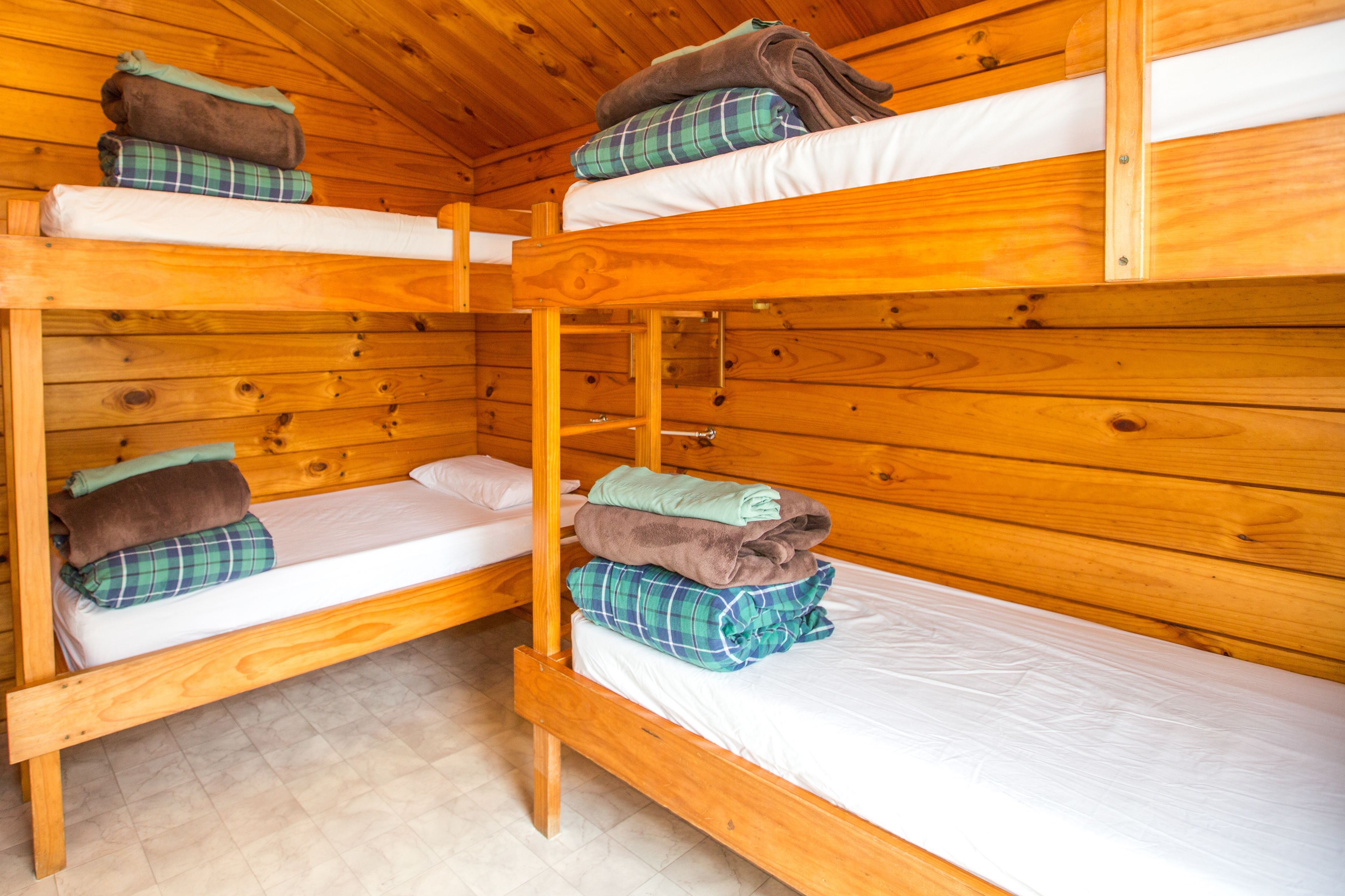 Quarto com beliches, paredes e chão de madeira, com duas camas equipadas com almofadas e cobertores.