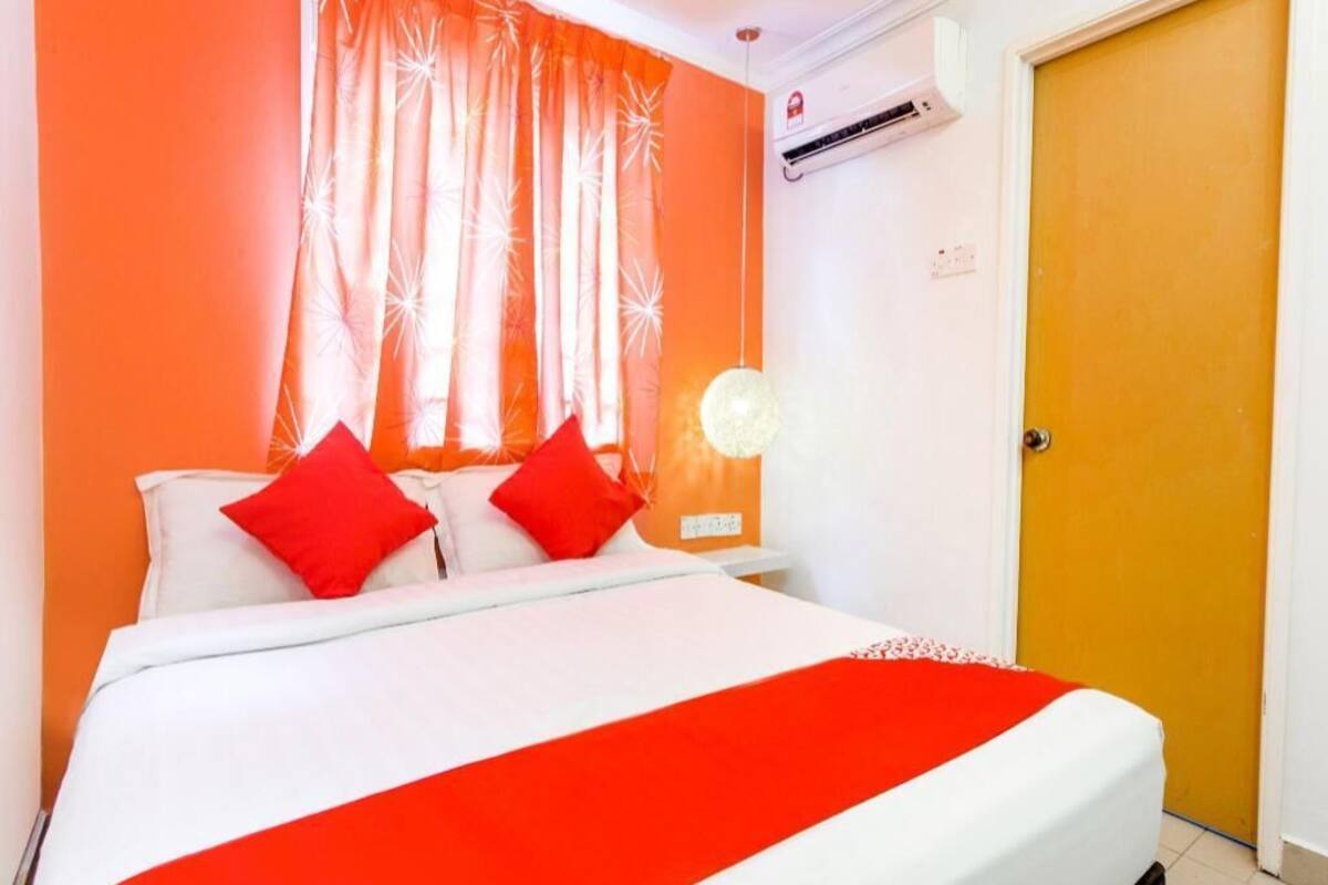 Quarto de hotel com cama, almofadas vermelhas, unidade de ar condicionado, porta e luminária fixada na parede.