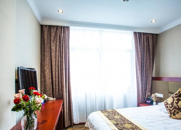 Quarto de hotel com cama, escrivaninha com um vaso de flores, televisão e cortinas.