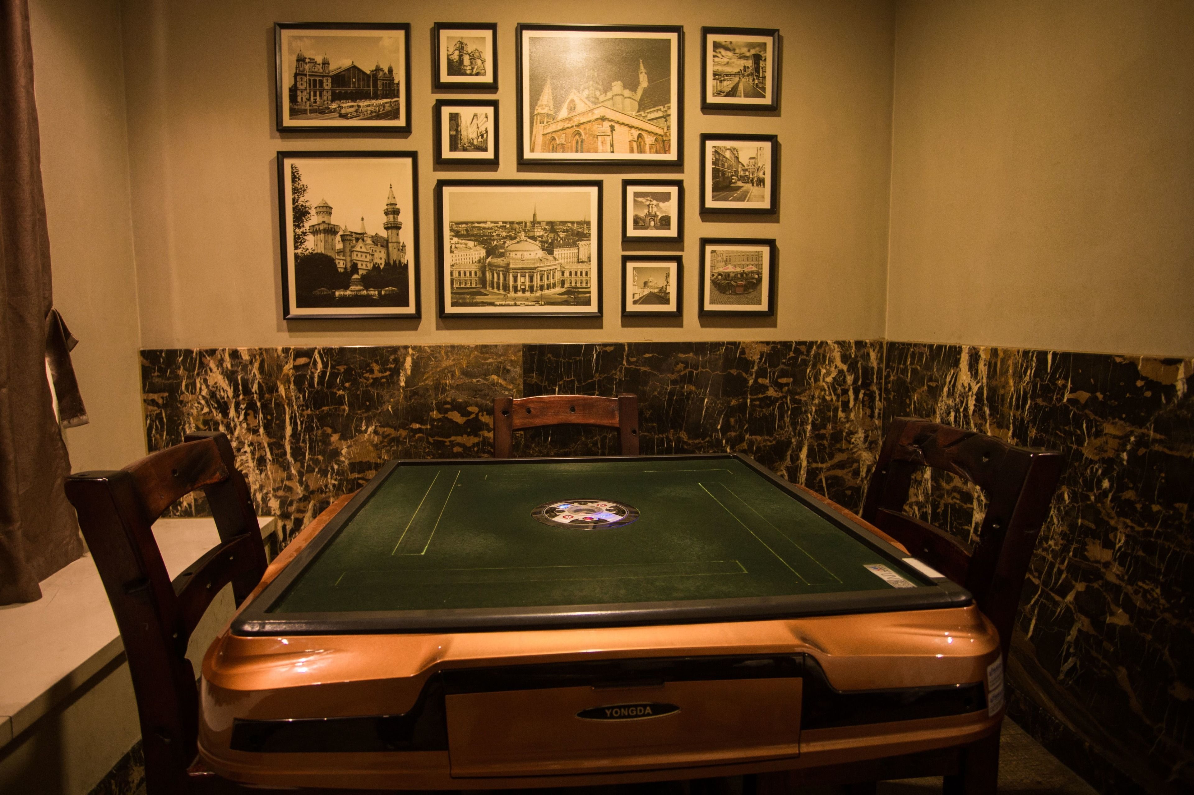 Uma mesa de bilhar com pano verde, rodeada por cadeiras de madeira. Acima da mesa, há quadros emoldurados na parede.