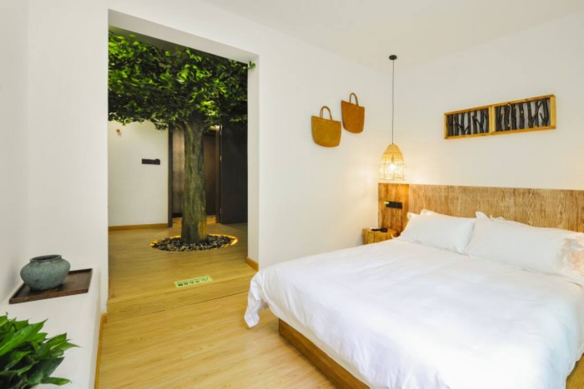 Quarto com cama, mesa de cabeceira, luminária na parede, cabeceira de madeira e uma árvore ao fundo.