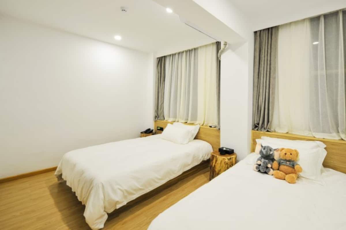 Quarto de hotel com duas camas de solteiro, piso de madeira, mesa de cabeceira com abajur e janela com cortinas.