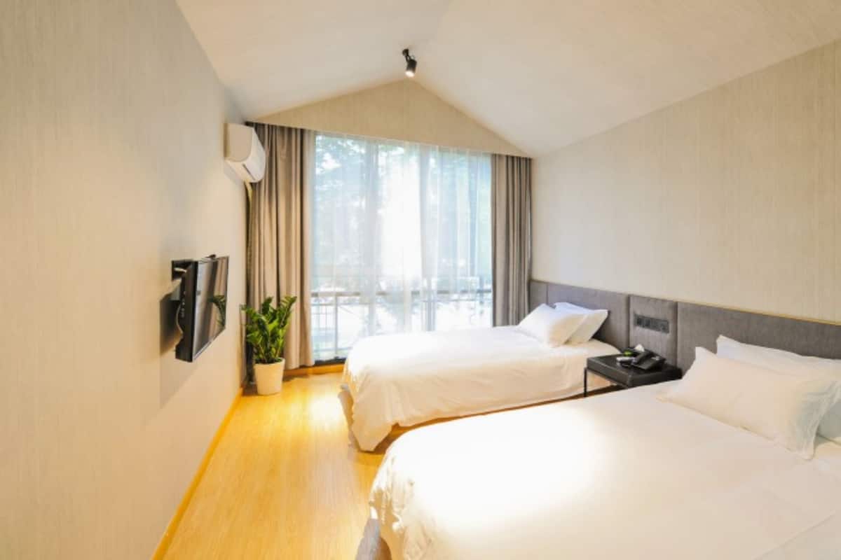 Quarto de hotel com duas camas, televisão na parede, planta em vaso e uma janela grande com cortinas.