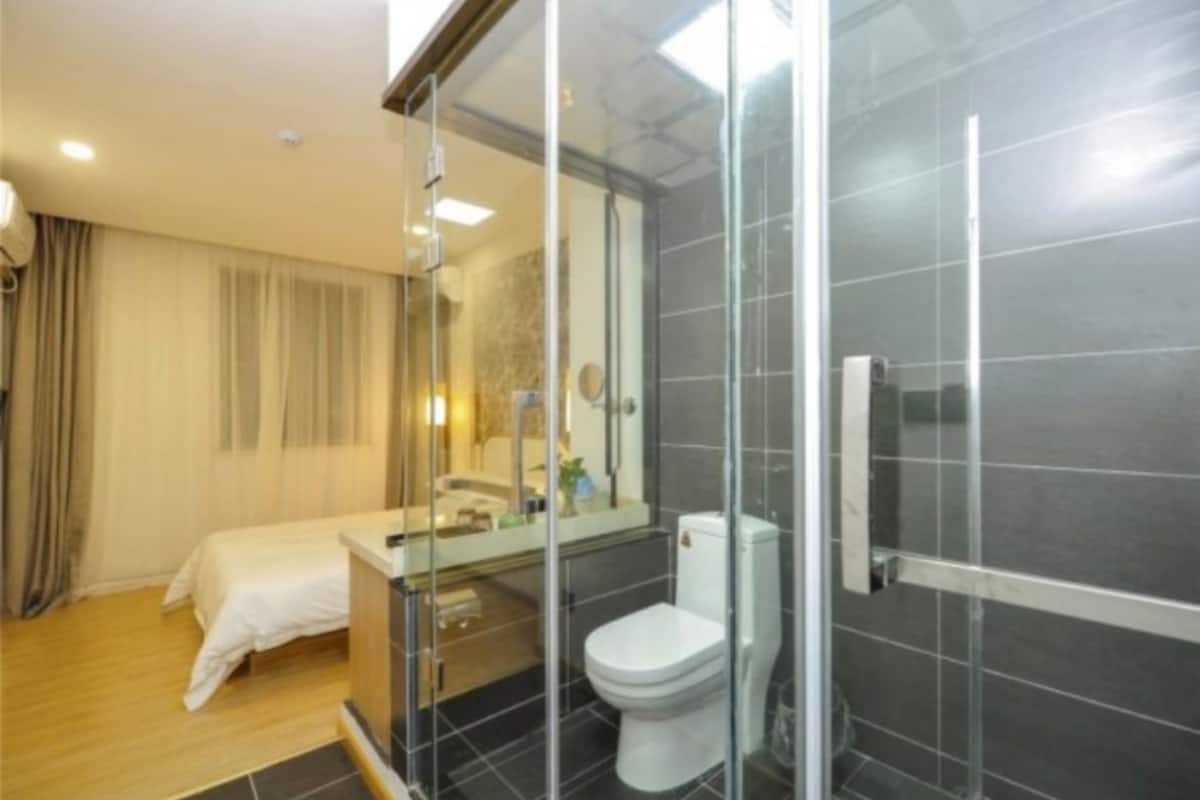 Quarto de hotel com chuveiro em cabine de vidro, cama, mesa de cabeceira e vaso sanitário.