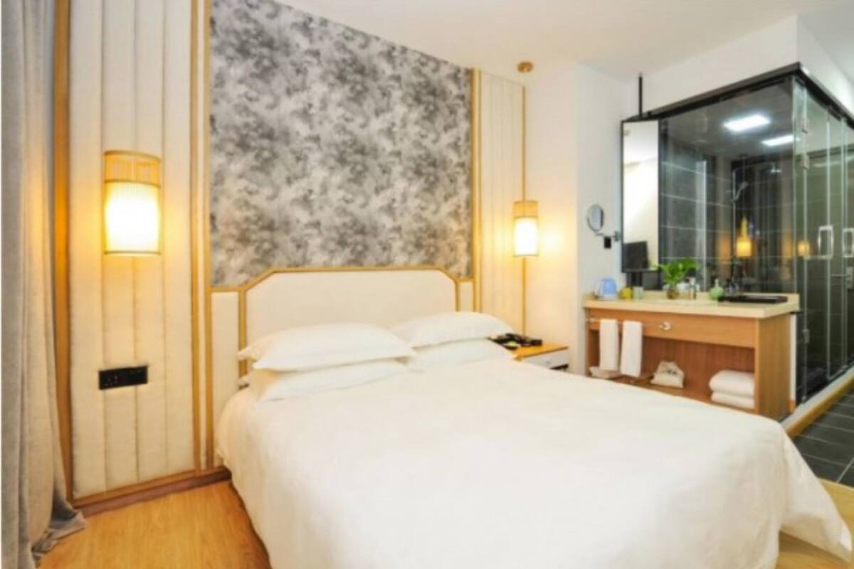Quarto de hotel com cama, luminárias de cabeceira, espelho e banheiro com box de vidro.