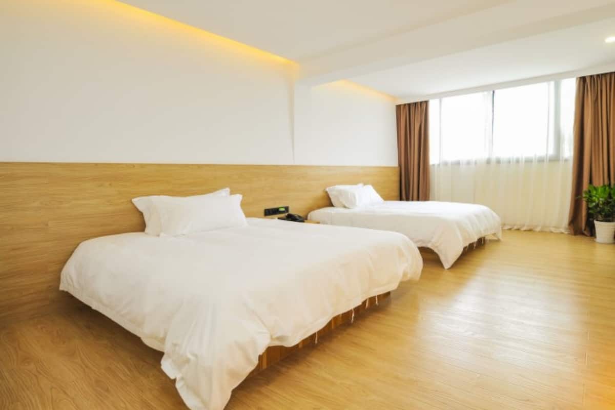 Quarto de hotel com duas camas, cabeceiras de madeira, roupa de cama branca, uma planta em vaso e uma janela com cortinas.