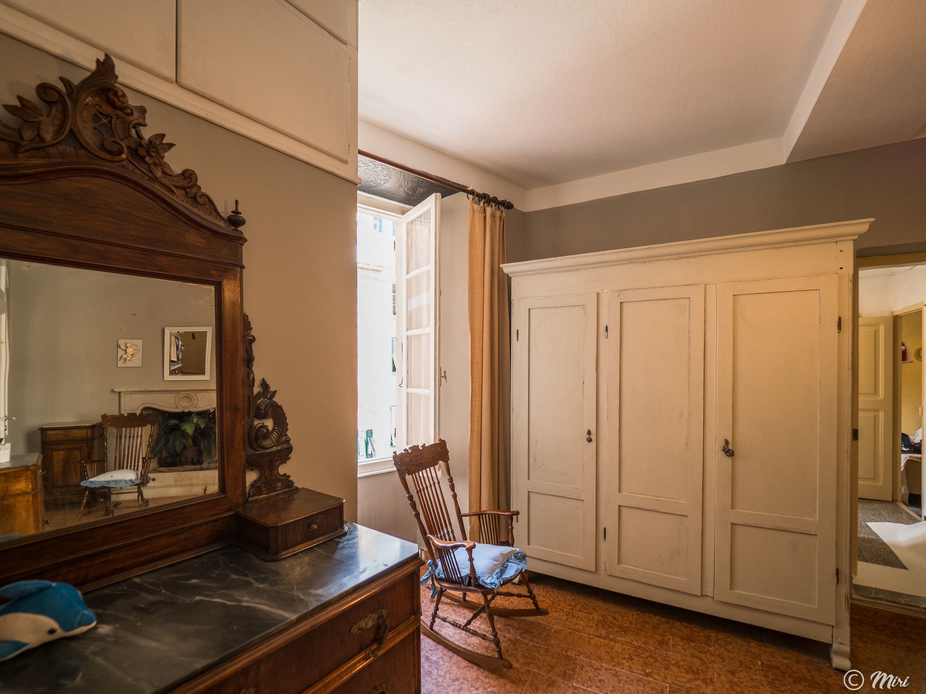 Quarto com cómoda de madeira, uma cadeira de balanço e um guarda-roupa grande e branco.