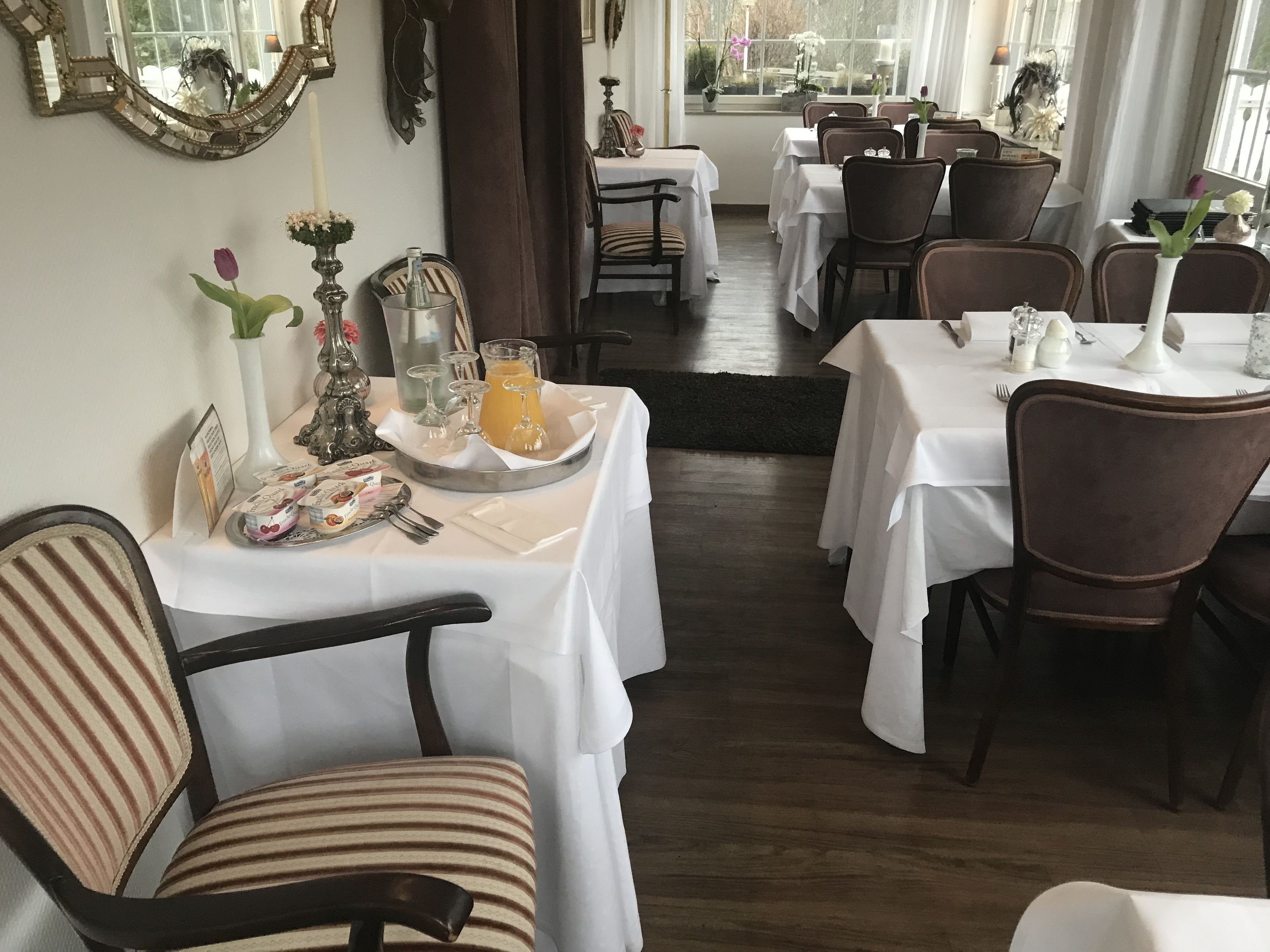Uma área de refeições com mesas preparadas para o serviço, um buffet com comida e bebidas, e vista para o exterior através de grandes janelas.