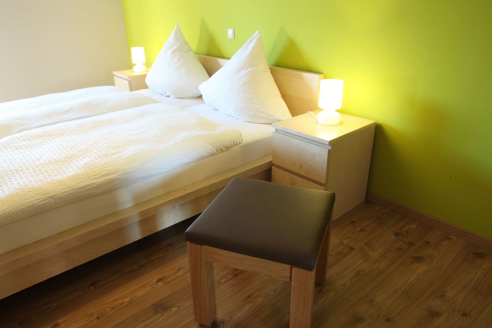 Cama bem arrumada com travesseiros e mesa de cabeceira com abajur, em quarto com piso de madeira e parede verde.