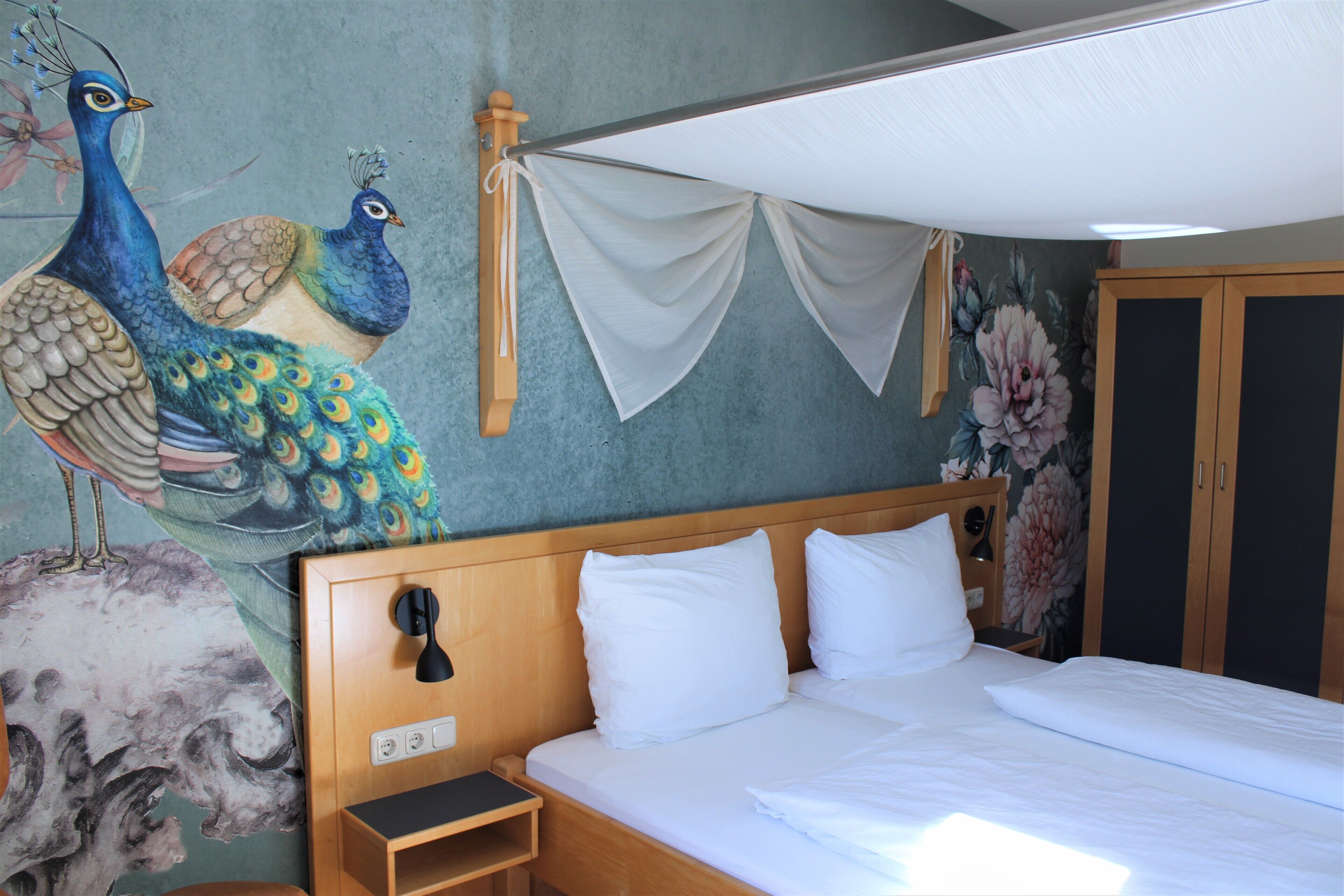 Quarto com cabeceira de madeira, cama com dossel, mesa de cabeceira e um mural de pavões na parede.