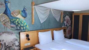 Quarto com cabeceira de madeira, cama com dossel, mesa de cabeceira e um mural de pavões na parede.