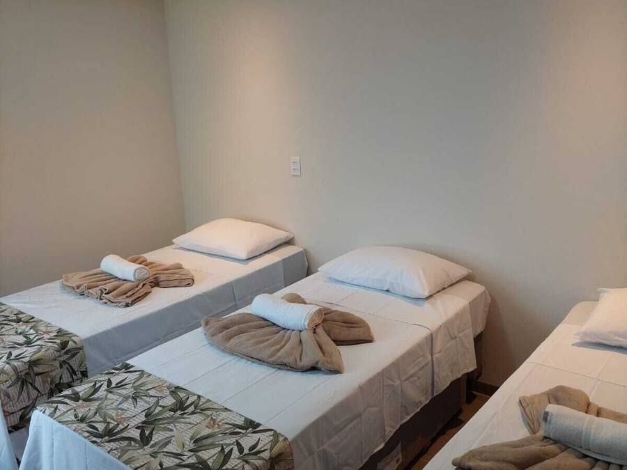 Quarto de hotel com três camas de solteiro, cada uma com uma almofada branca e uma toalha castanha. O quarto tem uma disposição simples, com paredes lisas e uma lâmpada no teto.