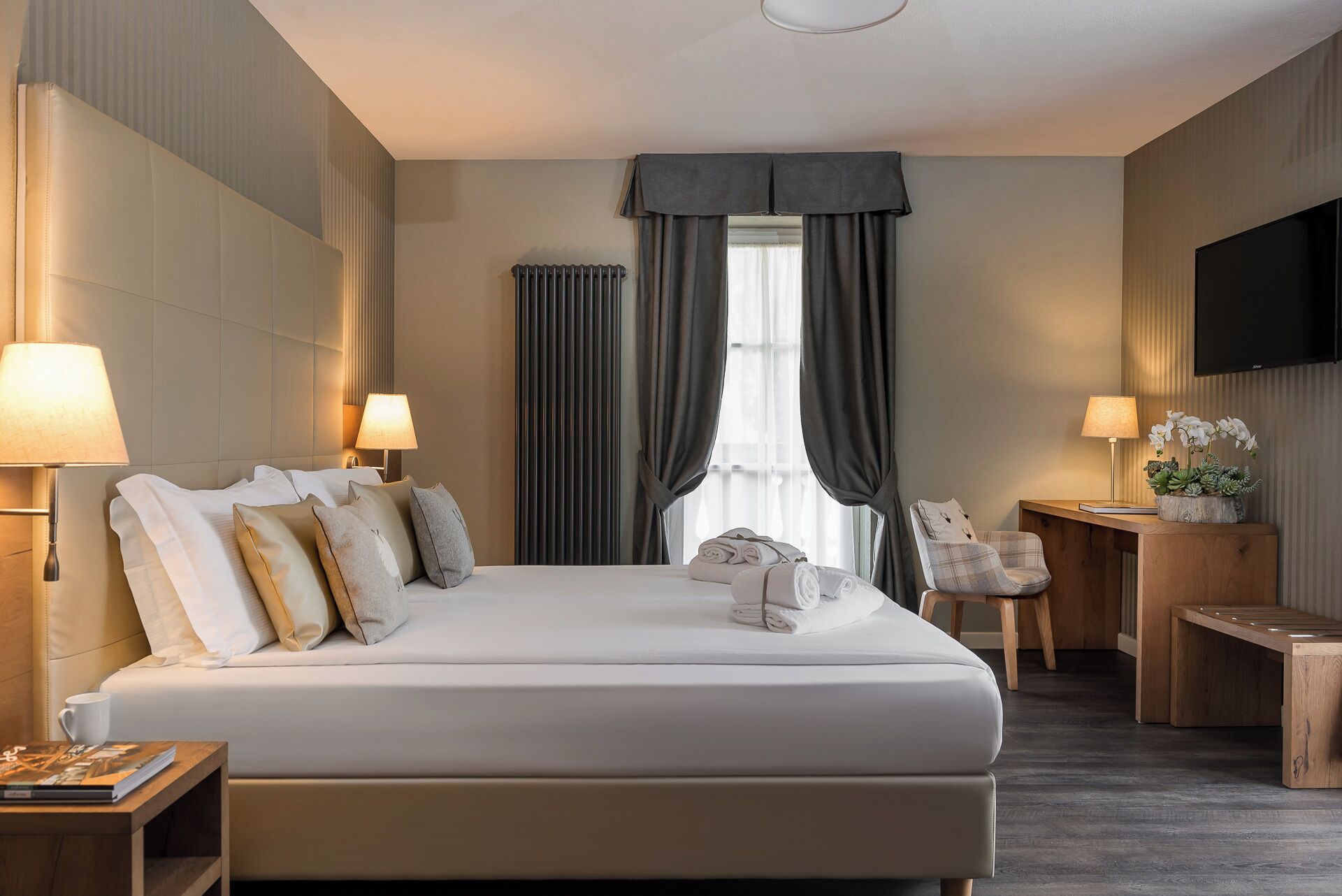 Quarto de hotel com uma cama grande, luminárias de cabeceira, uma mesinha com uma planta e uma televisão na parede.