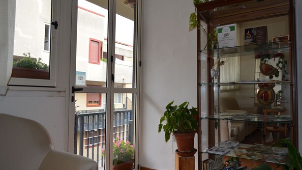 Quarto com porta de vidro deslizante que dá para uma varanda, estante cheia de livros e objetos decorativos, e um vaso com planta.