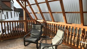 Varanda com duas cadeiras, uma mesa e balaústres de madeira, com vista para edifícios vizinhos.