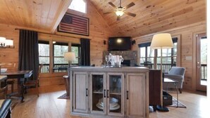 Interior de cabana com teto de madeira, área de refeições, cozinha com ilha central e televisão fixada na parede.