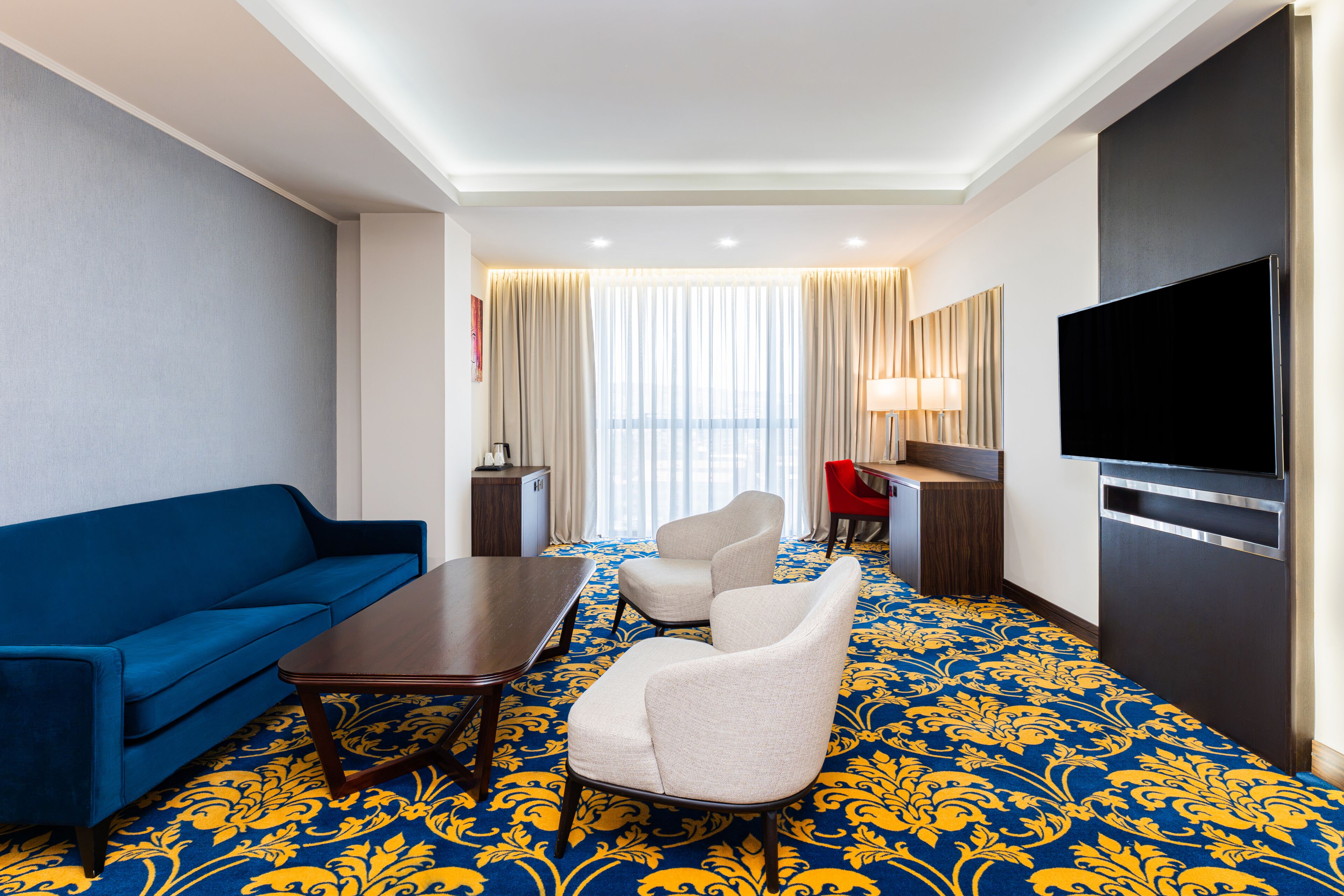 Quarto de hotel com um sofá azul, uma mesa de jantar, duas poltronas, uma televisão e uma janela grande com cortinas.