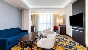 Quarto de hotel com um sofá azul, uma mesa de jantar, duas poltronas, uma televisão e uma janela grande com cortinas.