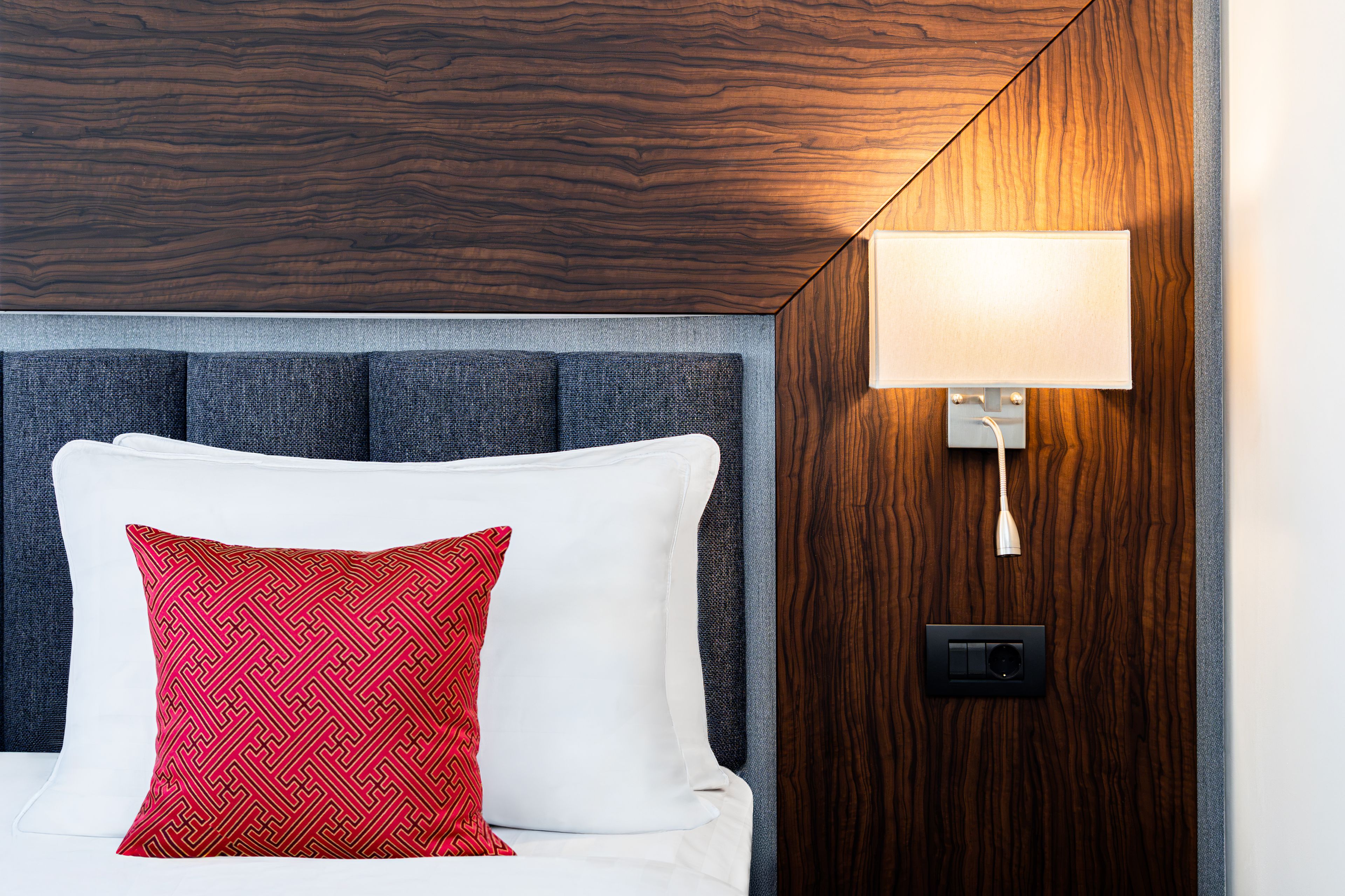 Quarto de hotel com cama, travesseiro estampado vermelho, cabeceira cinza e luminária fixada na parede.