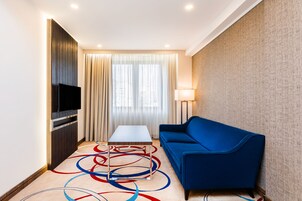 Quarto de hotel com televisão de tela plana, sofá azul, mesa pequena e luminária de chão.