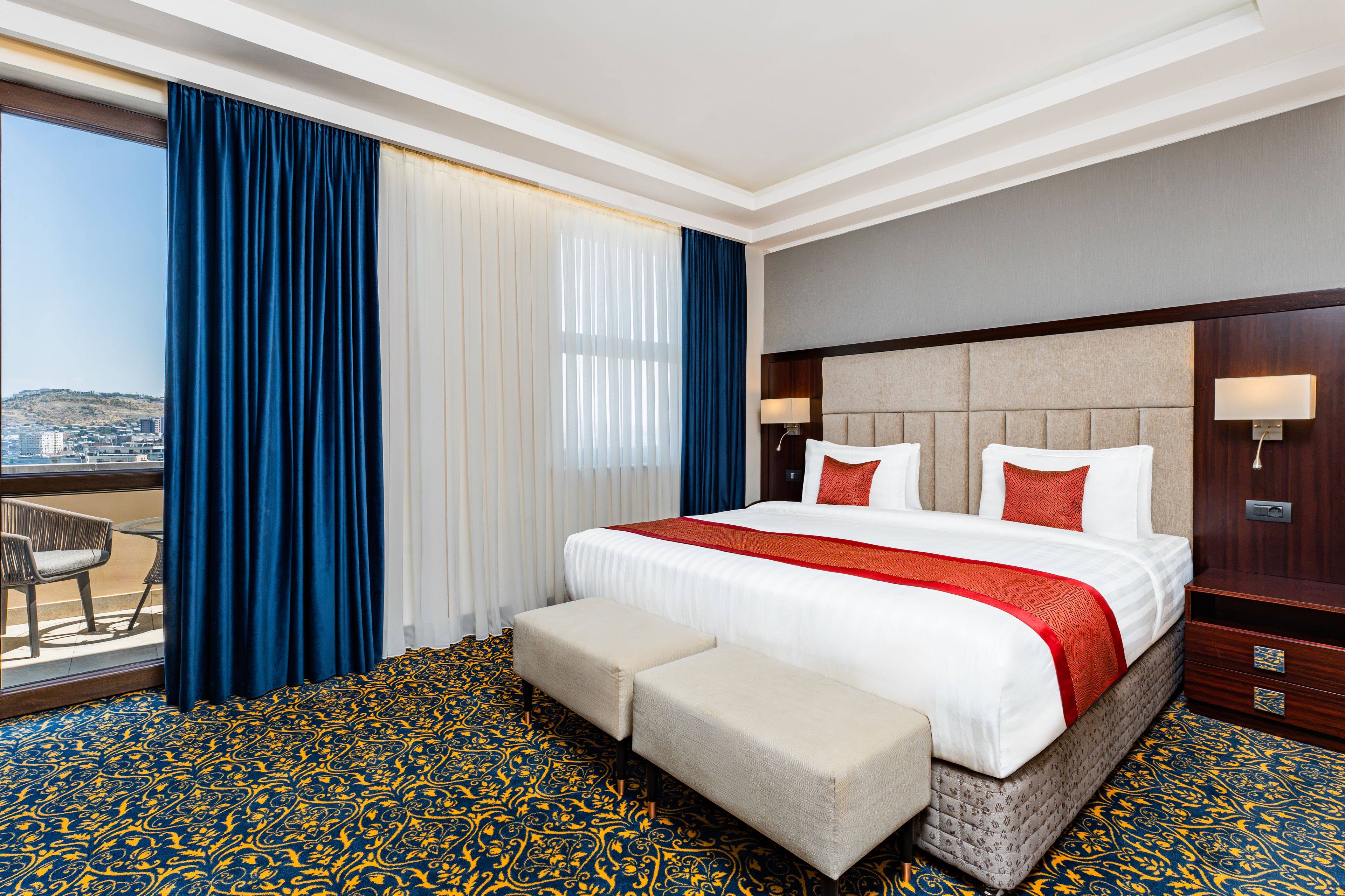 Quarto de hotel com uma cama grande, uma varanda com cortinas azuis e um tapete estampado.